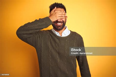 녹색 스웨터와 셔츠를 입은 젊은 인도 남자가 고립 된 노란색 배경 위에 서서 미소를 짓고 얼굴을 가리는 눈으로 웃고 있습니다