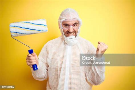 화가 장비를 착용하고 고립 된 노란색 배경 위에 그림 롤러를 들고 자랑스럽게 비명을 지르며 승리와 성공을 축하하는 매우 흥분되고