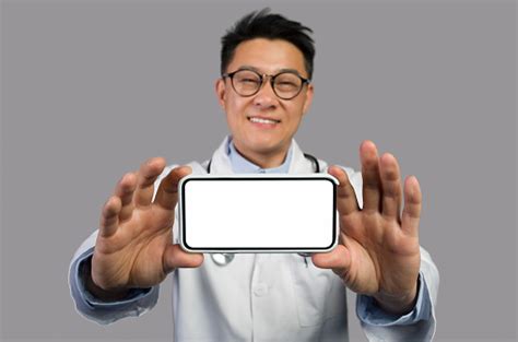 흰 코트와 안경을 쓴 기쁜 중년 아시아 남자 치료사는 빈 화면으로 스마트폰을 보여줍니다 35 39세에 대한 스톡 사진 및 기타