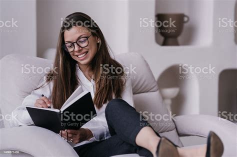 안경을 쓴 만족스러운 갈색 머리 사업가 아늑한 의자에 앉아있는 셔츠 검은 바지 열린 플래너가 활짝 웃으며 할 일 목록을