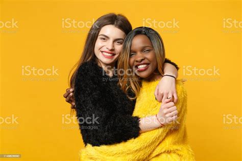 두 젊은 여자 친구는 밝은 오렌지 벽 배경 스튜디오 초상화에 고립 된 포즈를 서 있는 검은 노란 옷에 유럽과 아프리카계 미국인