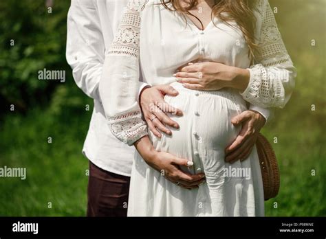 Pregnant woman and man posing at summer park. Close up of human hands holding pregnant belly ...