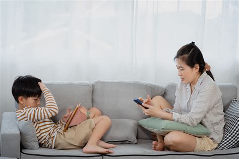 아시아계 미혼모가 집 거실의 소파에 앉아 태블릿을 사용하고 아들과 함께 옆에서 행복하게 게임을하고 있습니다 여자와 소년이 서로