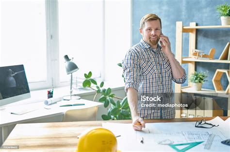 현대 디자인 스튜디오에서 작업 하는 동안 청사진 및 휴대 전화에 얘기 책상에 서 서 건설 회사의 쾌활 한 잘생긴 젊은 남성 관리자 건설 산업에 대한 스톡 사진 및 기타