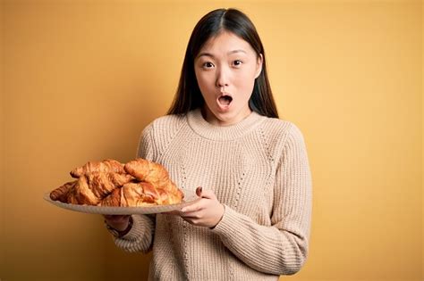 젊은 아시아 여자 들고 프랑스 과자 크루아상 위에 노란색 고립 된 배경 에 무서워 충격으로 충격 으로 깜짝 얼굴 두려움과 두려움