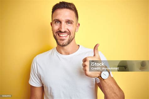 노란색 고립 된 배경 위에 캐주얼 흰색 티셔츠를 입고 젊은 잘 생긴 남자는 손으로 제스처를 행복 엄지 손가락을하고 성공을 보여주는 카메라를 보고 표정을 승인합니다 엄지
