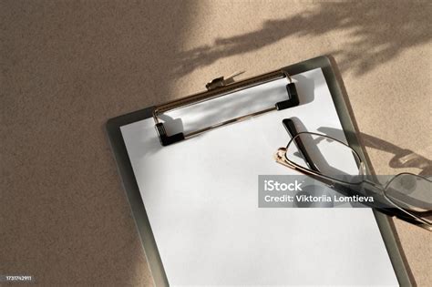 클립보드 모형에 있는 빈 종이 시트 미적 햇빛 그림자가 있는 베이지색 책상 배경에 있는 안경 평평한 위치 복사 공간 우아한 비즈니스 브랜딩 템플릿 0명에 대한 스톡 사진 및