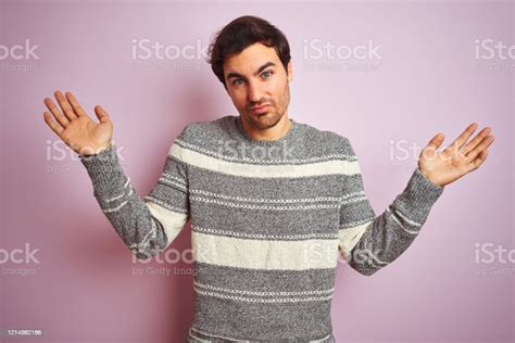 캐주얼 스트라이프 스웨터를 입은 젊은 잘생긴 남자는 고립 된 분홍색 배경 에 서 단서와 팔과 손을 들고 혼란 스러운 표정 의심