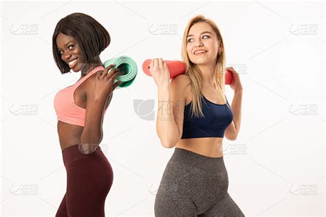 非洲和欧洲的运动女孩穿着运动服背靠背站着，国际女孩拿着瑜伽垫在她们的肩膀上孤立在白色的背景