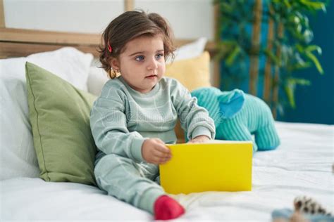 Adorable Hispanic Girl Reading Book Sitting On Bed At Bedroom Stock