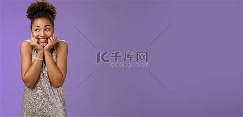 穿着银色裙子的傻傻可爱的黑人女孩期待着激动人心的派对梦幻般的凝视触摸脸颊微笑着广泛乐观等待梦想成真站立高兴触摸蓝色背景高清摄影大图 千库网