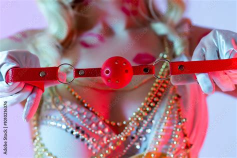 Beautiful Blonde In A White Sexy Dress And White Gloves Holds A Red Gag For Bdsm In Her Hand