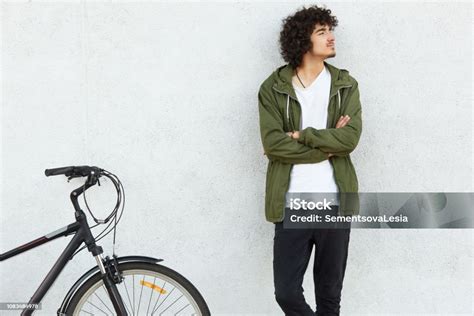 매력적인 남자의 실내 샷 손에 넘어 유행 옷 자전거 화이트 스튜디오 벽에 절연 근처 스탠드 입고 신중 하 게 보인다 사람 사이클링 및 생각 개념 곱슬 머리에 대한 스톡 사진