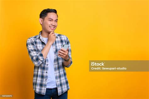 격자 무늬 셔츠를 입은 쾌활한 젊은 아시아 남자가 휴대 전화를 들고 노란색 배경에 고립 된 복사 공간을 옆으로 바라 봅니다 30 34세에 대한 스톡 사진 및 기타 이미지