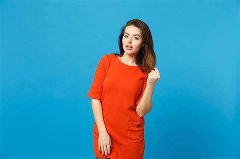 아름 다운 갈색 머리 젊은 여 자가 레드 오렌지 드레스를 입고 카메라 서 있는 포즈 유행 블루 벽 배경 스튜디오 초상화에 고립
