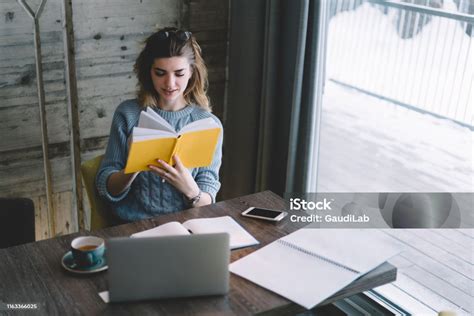 대학 캠퍼스에서 책과 함께 앉아 미소 여자 자유 시간 동안 흥미로운 문학을 즐기는 Autodidact에 시간을 보내는 성공적인 여성 학생 테이블에 머물고 현대 노트북 컴퓨터