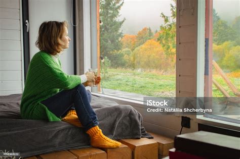 멋진 가을 풍경과 함께 큰 창문 뒤에서 차 한 잔과 함께 침대에 앉아있는 소녀 녹색 스웨터를 입은 여자가 안개 속에서 가을 산을 배경으로 하고 있다 산장에서 탈출하세요 집에