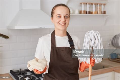 흰색 티셔츠와 갈색 앞치마를 입고 검은 머리를 한 웃는 여성의 실내 샷 손에 걸레와 천을 들고 가정부는 행복한 표정으로 카메라를 보고 있습니다 30 34세에 대한 스톡 사진