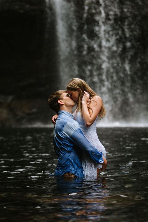 A Man And Woman Are Kissing In Front Of A Waterfall While Standing In