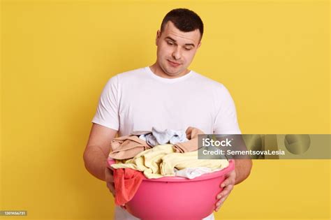 더러운 닫기로 장미 분지를 들고 피곤 한 잘 생긴 유럽 남자는 실망 얼굴 표정으로 린넨을보고 슬픈 노란색 벽에 서 많은 린넨을 세탁해야합니다 25 29세에 대한 스톡 사진