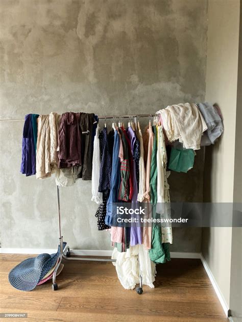 질감이 있는 회색 벽에 다양한 색상과 직물의 다양한 옷이 진열된 옷걸이 챙이 넓은 모자가 나무 바닥에 놓여 있습니다 0명에 대한