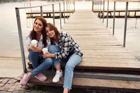 Cute Lesbian Couple Sits Together Near The Lake With Cups Of Drink In