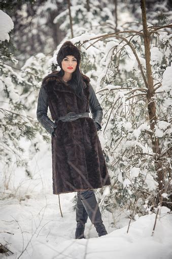 긴 검은 모피 코트와 숲에서 겨울 풍경을 즐기는 모자에 아름 다운 여자 눈 덮인 나무 아래에서 포즈를 취한 갈색 머리 소녀 숲에서 분기 밝은 추운 날에 매력적인 젊은 여자