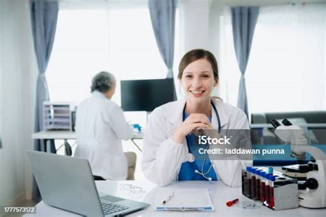 흰 의료복을 입은 자신감 넘치는 젊은 백인 여성 의사가 책상에 앉아 컴퓨터 작업을 하고 있다 웃는 사용 노트북 의학 저널에 쓰기 25 29세에 대한 스톡 사진 및 기타