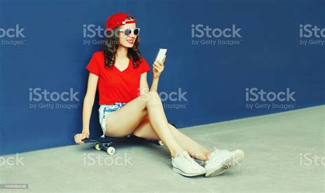 빨간 야구 모자를 쓰고 스케이트 보드에 스마트 폰을 들고 행복한 미소 짓는 젊은 여성 푸른 벽 배경에 도시 거리의 반바지 사람들에 대한 스톡 사진 및 기타 이미지 Istock