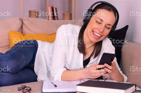 예쁜 갈색 머리 젊은 여자는 헤드폰을 통해 음악을 듣고 집에서 소파에 뻗어 공부 하는 그녀의 스마트 폰으로 웃고 Mp3