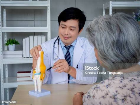 흰색 정장을 입은 전문 아시아 남성 정형외과 의사가 무릎 관절 해부학 모델을 가리키며 의료 사무실에서 고위 여성 환자를