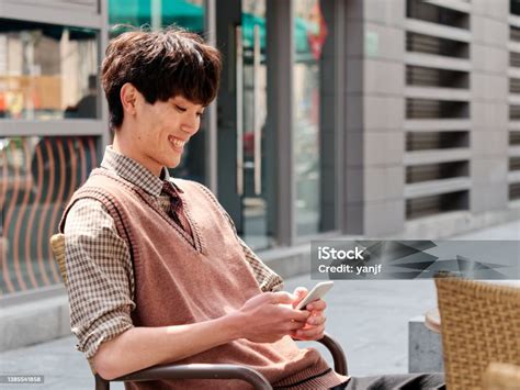 격자 무늬 셔츠와 울 조끼에 곱슬 검은 머리를 가진 잘 생긴 중국 청년의 초상화는 화창한 날에 손에 자신의 휴대 전화와 야외