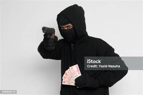 검은 후드티와 마스크를 쓴 수수께끼의 남자가 십만 루피아 지폐를 들고 총을 들고 총을 쏘며 돈을 갈취하는 초상화 회색 배경에