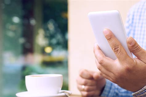 자른 이미지 젊은 남자 커피와 스마트 폰의 컵과 테이블에 앉아 전화에 메시지를 입력 하는 남자의 손을 터치 스크린 발코니 테라스에 앉아있는 동안 Brand Name