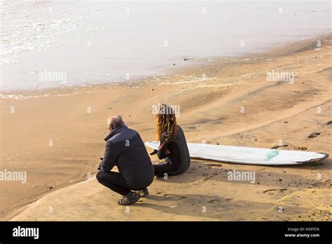 Surfer Sitting On The Beach Next To Surfboard With Another Man Looking