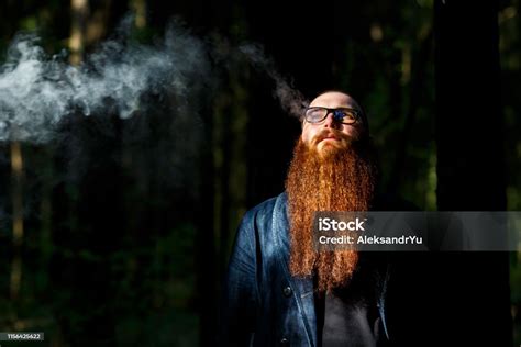 Vape 수염 남자 안경에 매우 긴 수염을 가진 성인 흰색 짧은 머리 남자는 어두운 숲에서 외부 전자 담배를 연기 나쁜 습관
