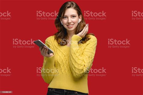 스튜디오에서 빨간색 배경에 전화 및 무선 이어폰을 가진 젊은 여성 그녀는 노란색 스웨터를 입고 설계를 위한 공간을 복사합니다 감정이 있는 개념 14에 대한 스톡 사진 및