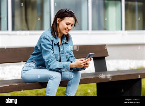 A Young Woman In Jeans And A Denim Jacket Sits On A Bench Smiling As