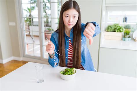 아름 다운 아시아 여자 먹는 녹색 신선한 브로콜리 화가 얼굴 부정적인 표시 혐오 아래로 엄지 손가락 거절 개념 가정 생활에 대한