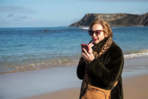 겨울 해변에서 립스틱을 바르고 휴대폰을 거울로 사용하고 코트와 선글라스를 착용한 노인 여성의 클로즈업 Brand Name Smart Phone에 대한 스톡 사진 및 기타