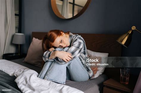 침대에 앉아 있는 우울한 밀레니얼 세대의 빨간 머리 여성의 앞모습은 나쁜 비관적인 생각에 휩싸인 무릎을 껴안고 혼자 고통받는다