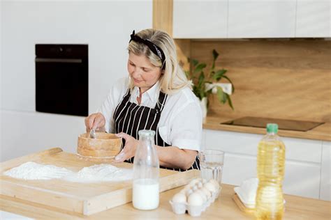 나무 테이블에 체질에 숟가락 가루를 붓는 체를 들고있는 행복한 노인 여성 현대 주방 베이킹에서 시간을 보내며 수제 케이크 피자 파스타를 요리하여 손주들을 놀라게합니다 가정