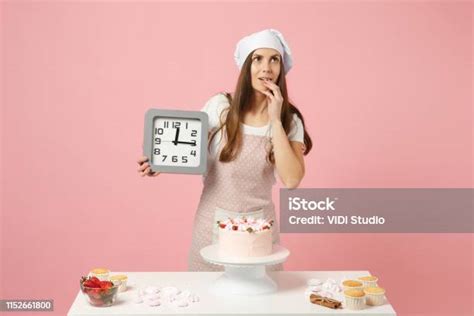 요리사 쿡 과자 또는 베이커 앞치마 흰색 T 셔츠 모자 요리사 모자 요리 케이크 또는 테이블 홀드 시계에서 분홍색 파스텔 배경에 고립 스튜디오에서 복사 공간 식품 시간