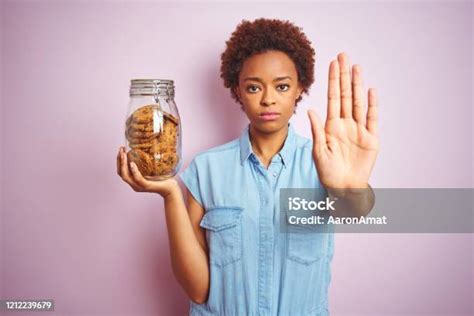 분홍색 고립 된 배경 위에 초콜릿 칩 쿠키 항아리를 들고있는 아프리카 계 미국인 여성은 진지하고 자신감있는 표정 방어 제스처로