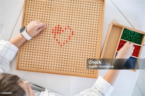 소년 아이는 책상에 빨간 구슬 하트 모양이 있는 곱셈 몬테소리 나무 보드를 사용합니다 목재 재료에 대한 스톡 사진 및 기타