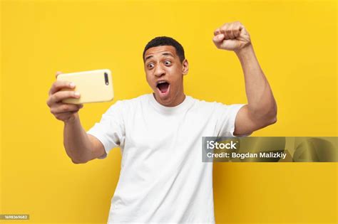 흰 티셔츠를 입은 젊은 아프리카계 미국인 남성이 스마트폰으로 모바일 게임을 하고 승리를 기뻐한다 비명에 대한 스톡 사진 및 기타 이미지 Istock