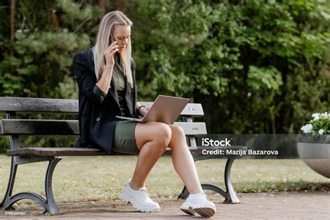 검은 블레이저를 입은 금발의 사업가가 여름 공원의 벤치에 앉아 노트북 컴퓨터에 타이핑하고 전화 통화를하고 몸짓을하고 야외에서 공부하고 있습니다 원격으로 일하는 여성 35