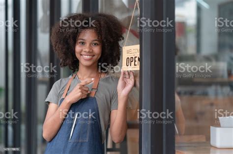 카페테리아 문 입구에 서 있는 아프리카계 미국인 사업가의 초상화 열린 간판이 있는 유리문 근처에 파란색 앞치마를 두르고 카메라를