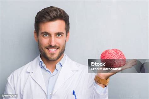 서 치아를 보여주는 자신감 미소로 웃 고 행복 한 얼굴로 뇌를 들고 회색 그런 지 벽에 잘생긴 젊은 의사 심리학자 남자 Mental Wellbeing에 대한 스톡 사진 및