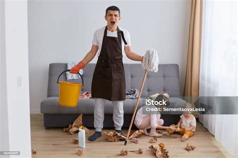 캐주얼 한 옷을 입고 앞치마를 씻고 아이들과 포즈를 취하고 걸레와 양동이를 손에 들고 비명을 지르며 충격을 받은 화가 난 남자의 실내 촬영이 엉망진창에 처했습니다 아버지에